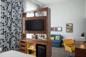 Quarto de hotel com uma estante de entretenimento de madeira, uma TV de tela plana, um refrigerador compacto, um sofá azul, uma poltrona cor mostarda e uma pequena mesa com abajur.