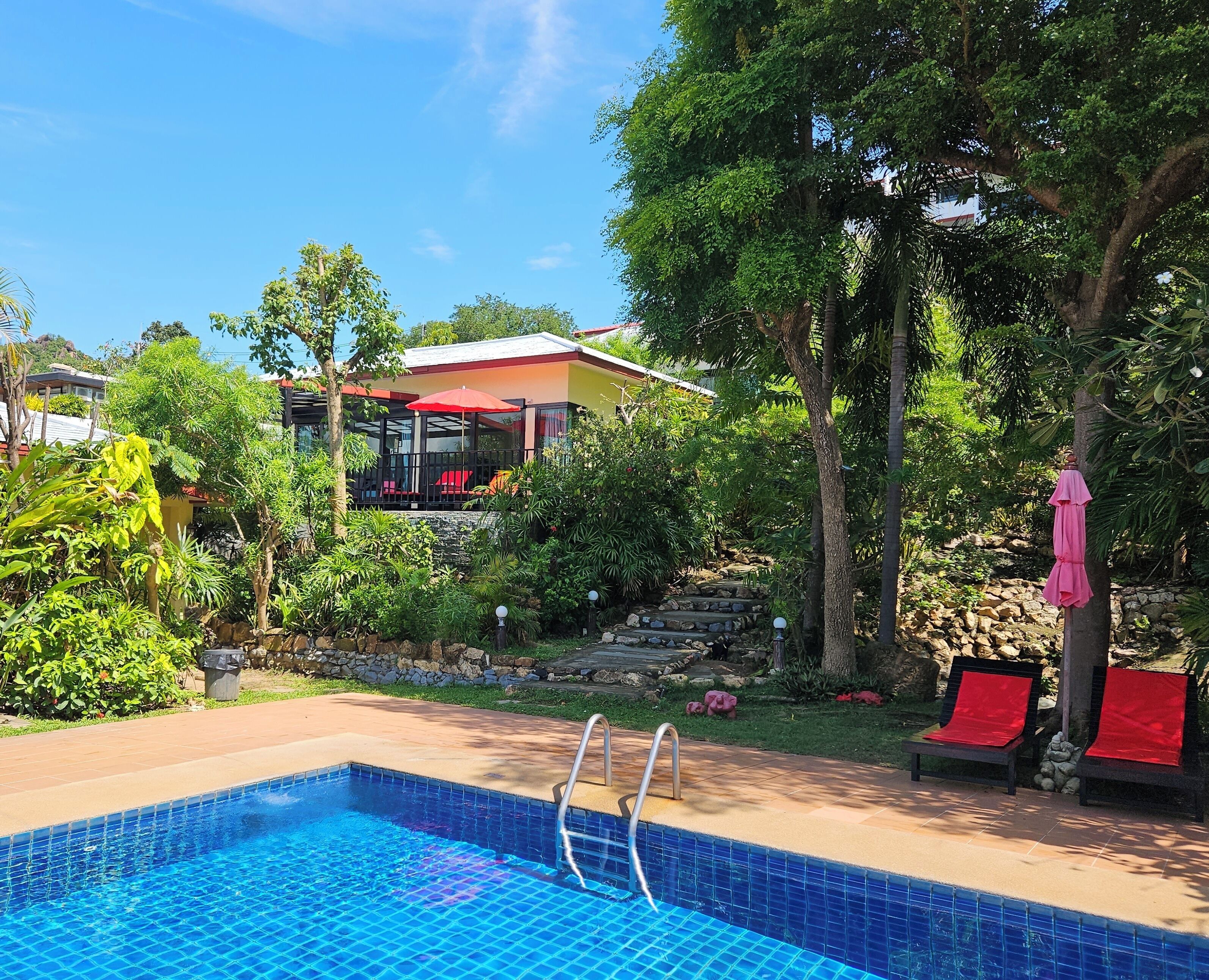 Uma piscina com borda azulejada, rodeada por vegetação e uma casa com um guarda-sol vermelho.