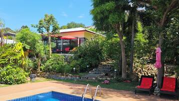 Uma piscina com borda azulejada, rodeada por vegetação e uma casa com um guarda-sol vermelho.