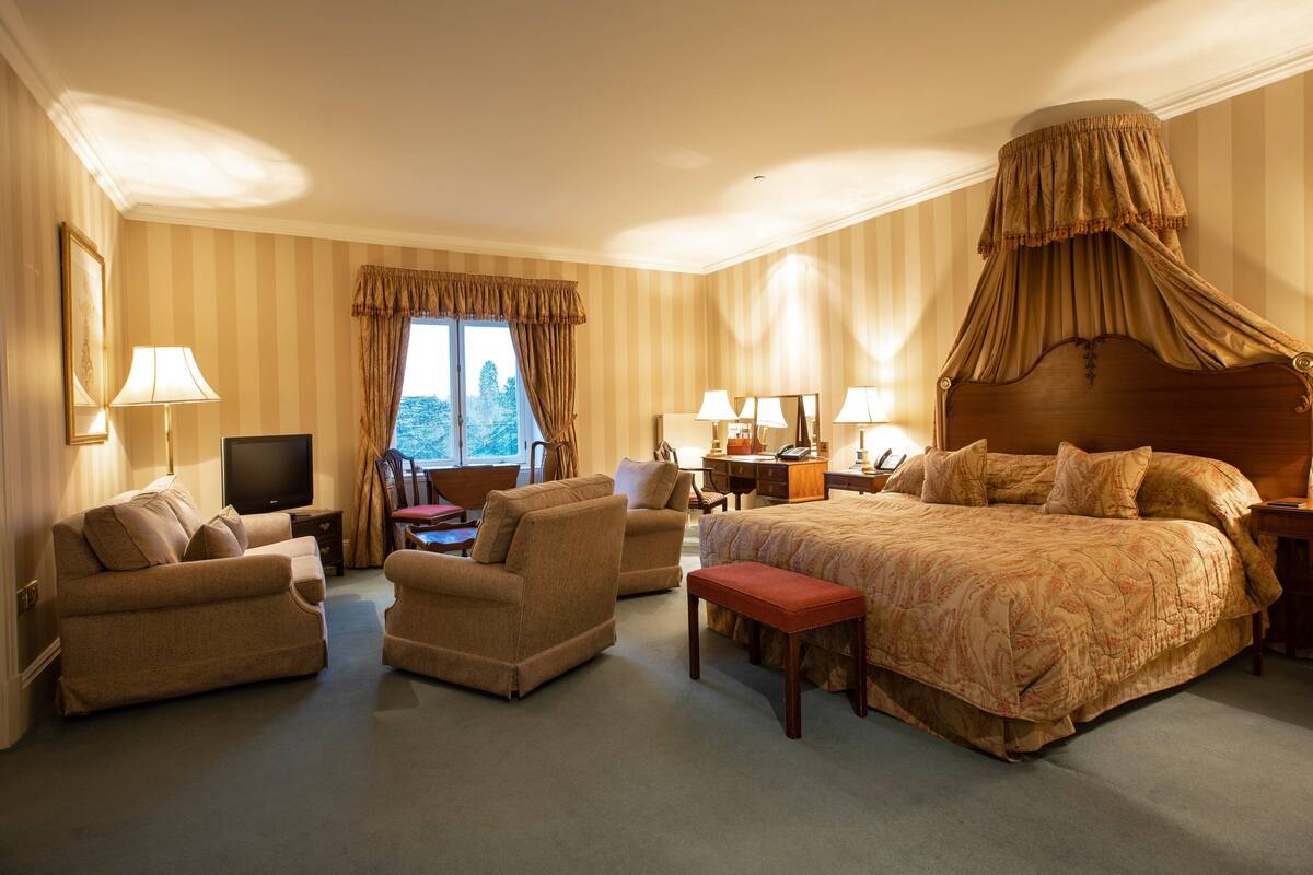 Quarto de hotel com uma cama grande, televisão, secretária e duas poltronas.