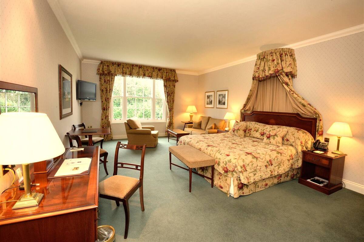 Quarto de hotel com uma cama grande, uma escrivaninha com luminária, uma cadeira, uma mesinha e uma televisão.