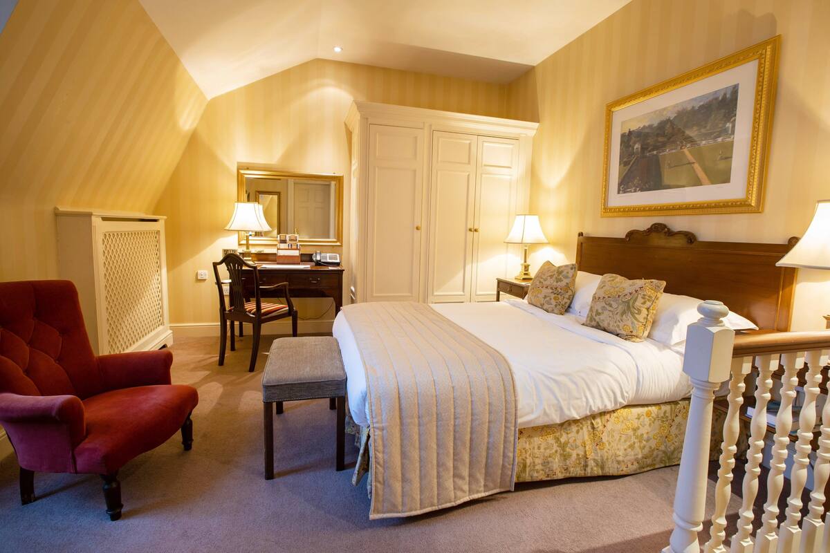 Quarto de hotel com uma cama grande, uma escrivaninha com cadeira, uma poltrona vermelha e um corrimão branco.