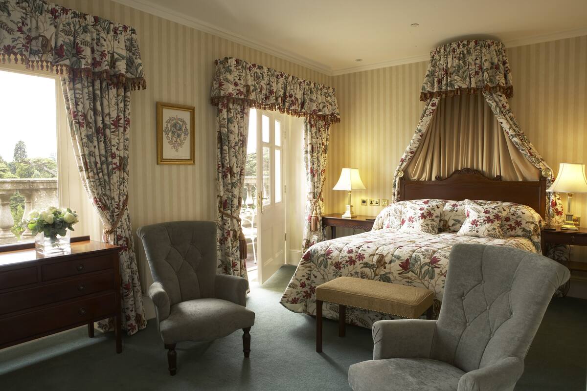 Quarto com uma cama grande, duas mesas de cabeceira com luminárias, uma cadeira, escrivaninha com um vaso de flores e uma janela com cortinas.