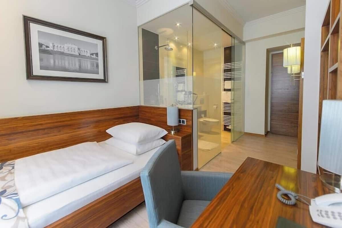 Quarto de hotel com cama, escrivaninha, cadeira, mesa de cabeceira, luminária, telefone, quadro na parede e porta de vidro para o banheiro.