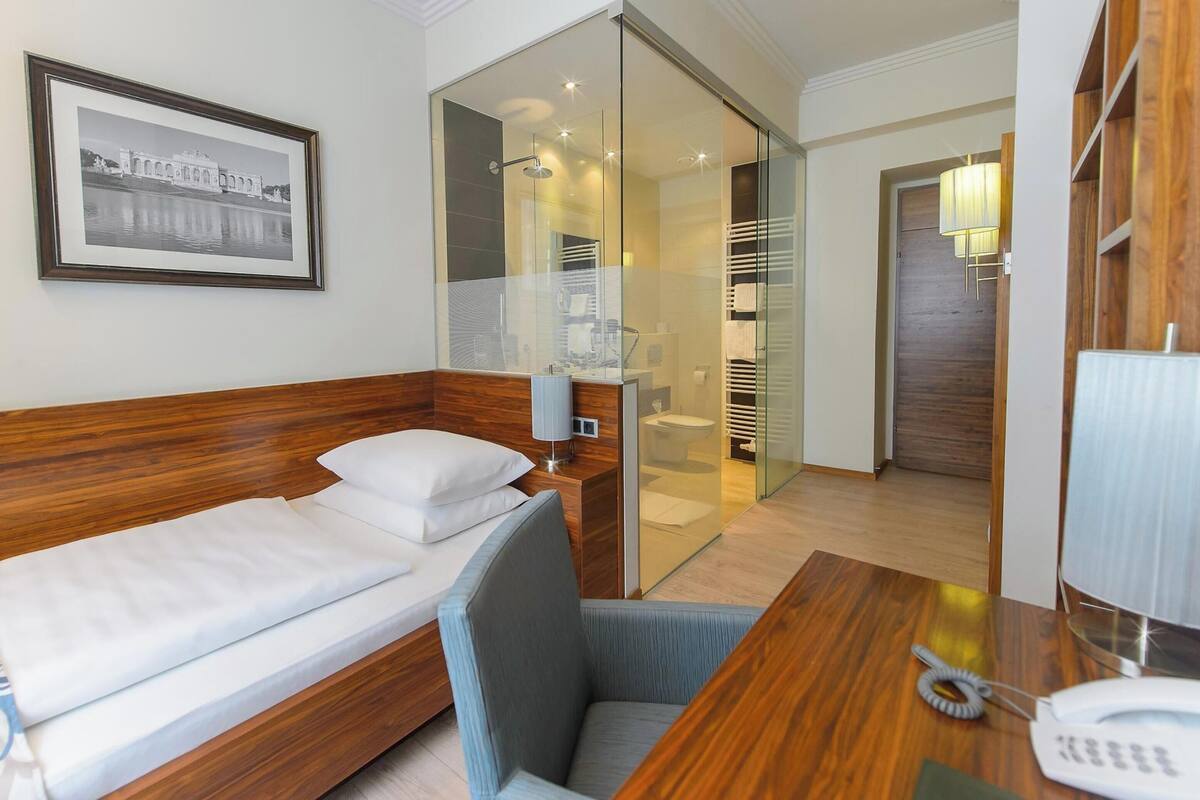 Quarto de hotel com cama de madeira, uma escrivaninha com telefone, uma cadeira, um painel de vidro e um quadro na parede.
