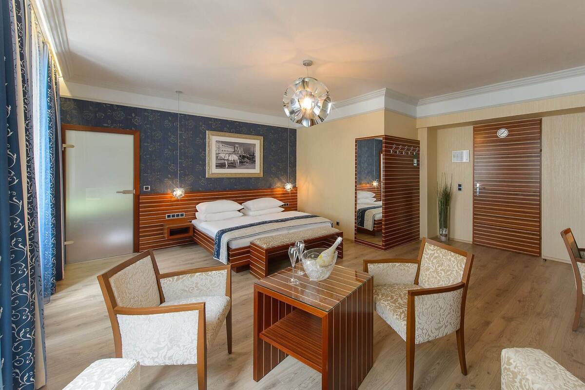 Quarto de hotel com uma cama grande, área de estar com mesa e cadeiras, espelho e lustre.