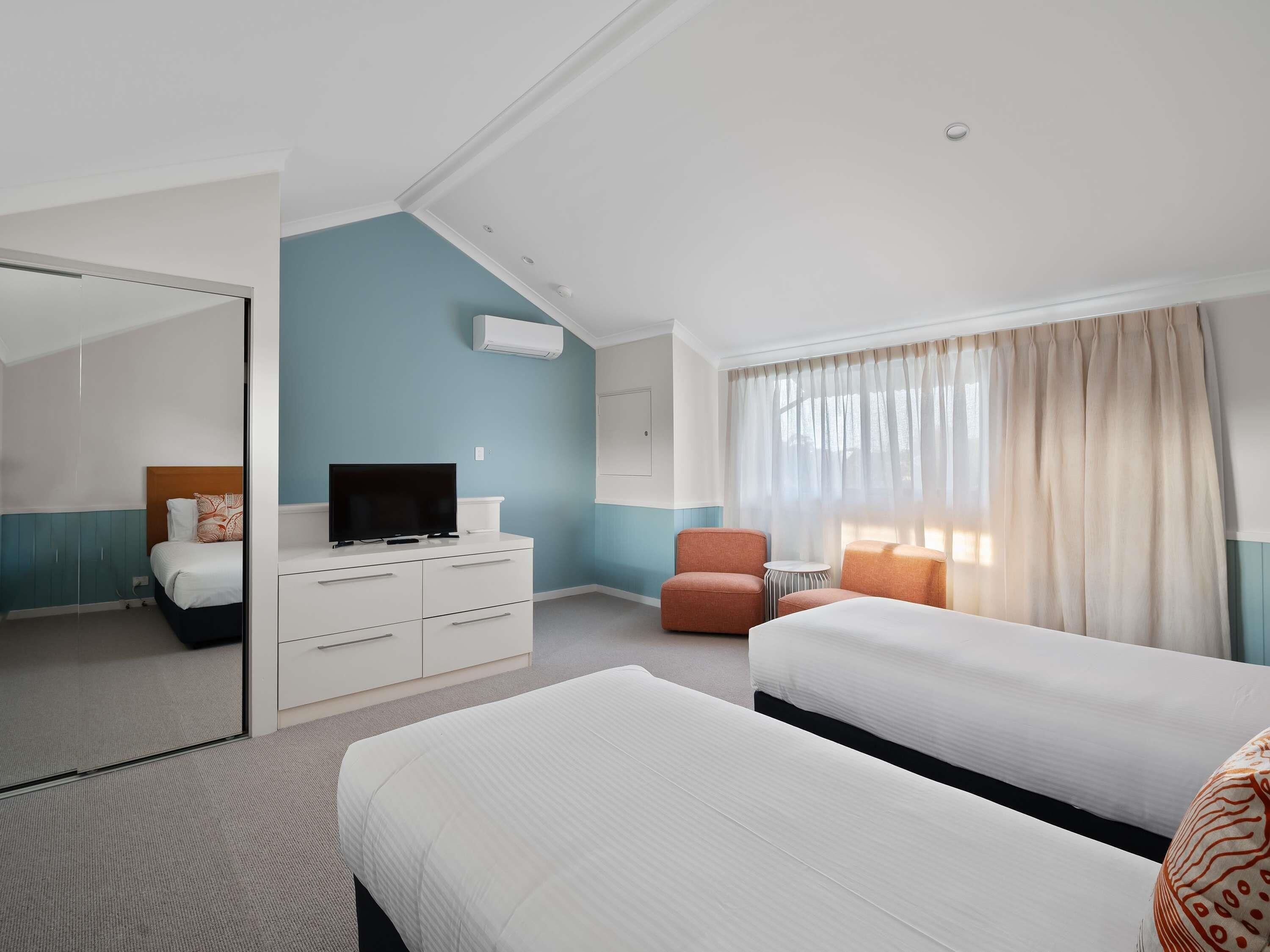 Quarto de hotel com uma cama, televisão sobre um aparador, uma cadeira e uma janela com cortinas.