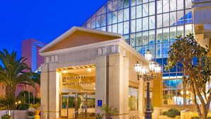 Entrada de hotel com fachada de vidro, uma placa com a inscrição "HOTEL" e área de acesso bem iluminada com plantas em vasos.