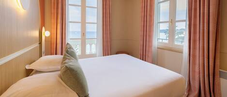 Quarto de hotel com cama, duas almofadas, mesa de cabeceira, luminária na parede e janela com cortinas.