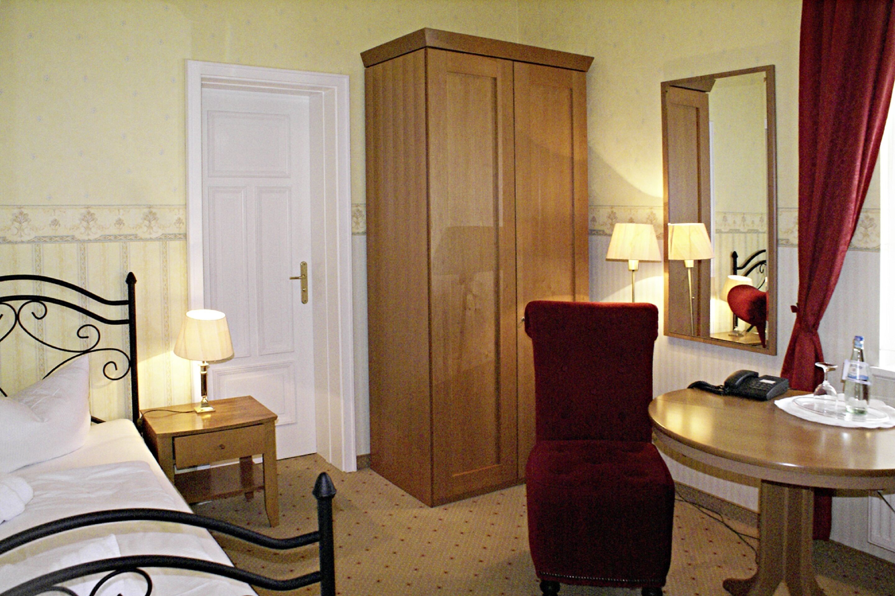 Quarto de hotel com cama, guarda-roupa de madeira, secretária com cadeira, mesa de cabeceira com luminária e espelho na parede.