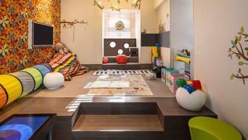 Uma sala de brinquedos com parede colorida, tapete de atividades, brinquedos e uma janela com persianas.