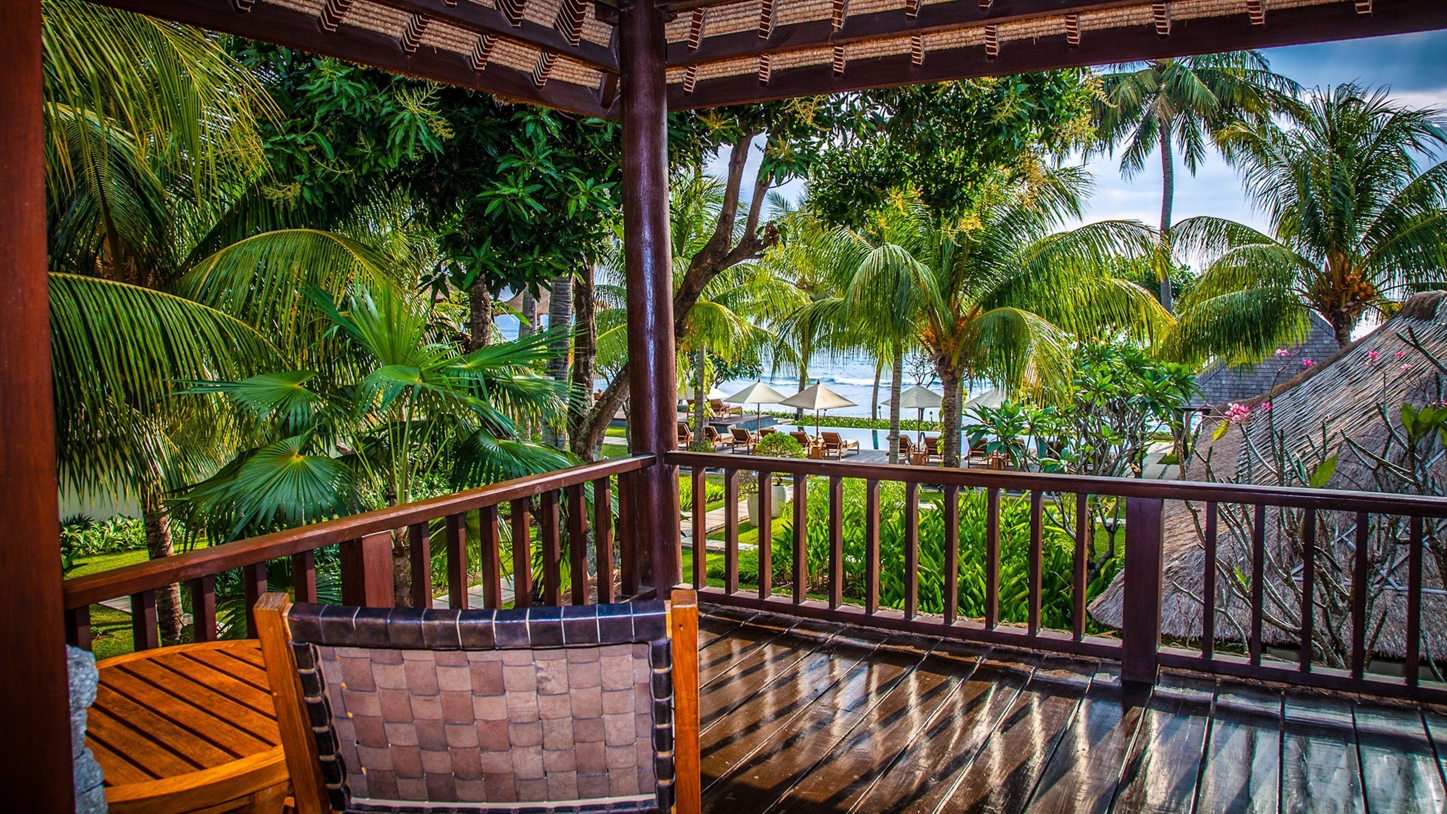Um deck de madeira com um banco revestido de azulejos, cercado por vegetação tropical exuberante e com vista para uma área de piscina.