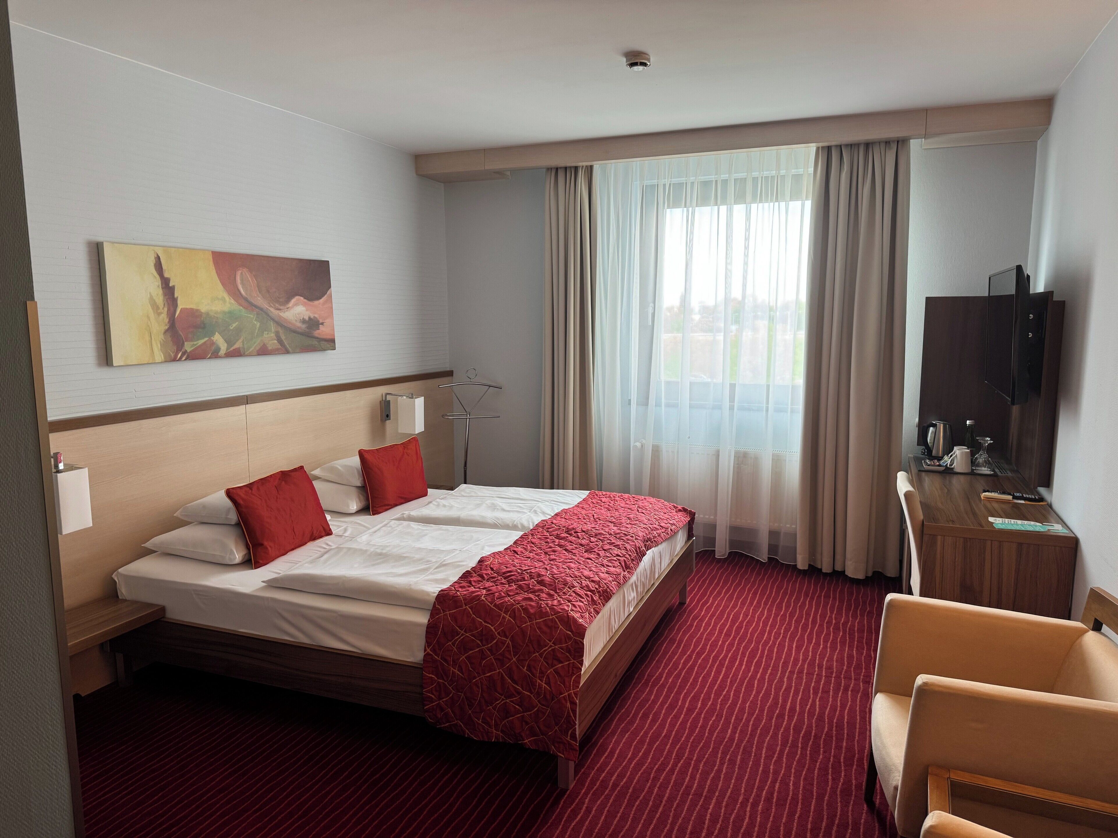 Quarto de hotel com uma cama grande, uma escrivaninha com cadeira, televisão e janela com cortinas.