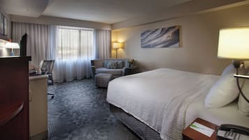 Quarto de hotel com uma cama grande, uma escrivaninha com luminária, uma cadeira, uma pequena mesa com um vaso e um quadro na parede.