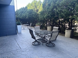 Um pátio com mesa e cadeiras, cercado por um sebedo e um edifício.