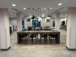 Uma sala de jantar espaçosa com uma mesa grande e cadeiras, iluminação pendente no teto e vista para o exterior através de amplas janelas.