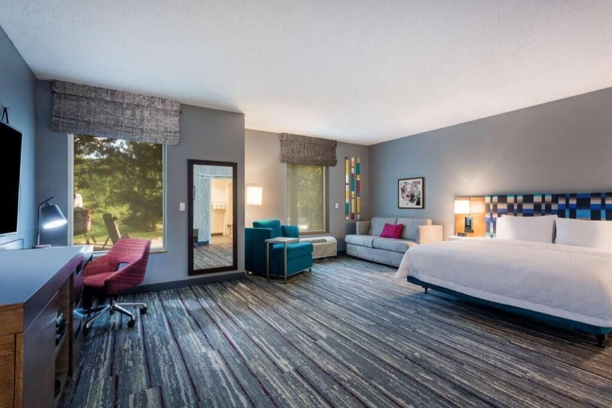 Quarto de hotel com cama, escrivaninha, cadeira, sofá e uma grande janela.