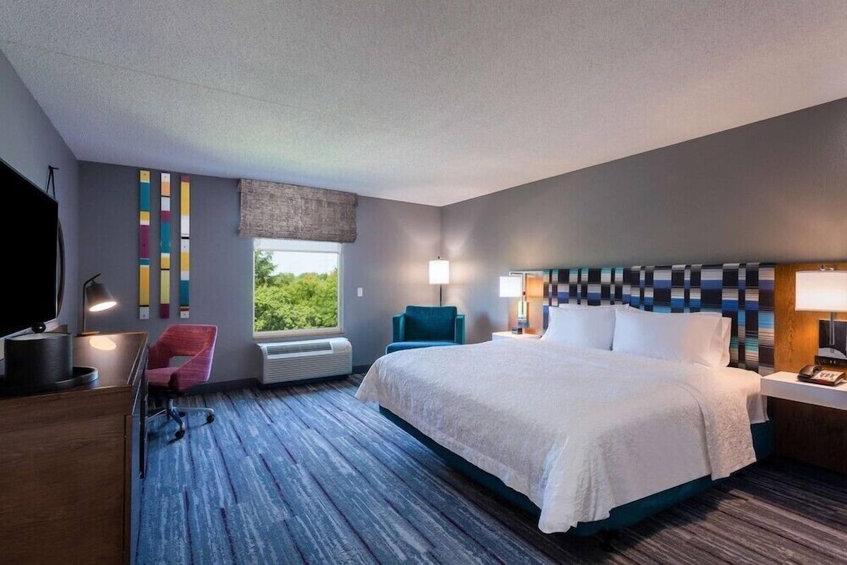 Quarto de hotel moderno com uma cama grande, uma mesa, uma cadeira, uma televisão e uma janela com vista.