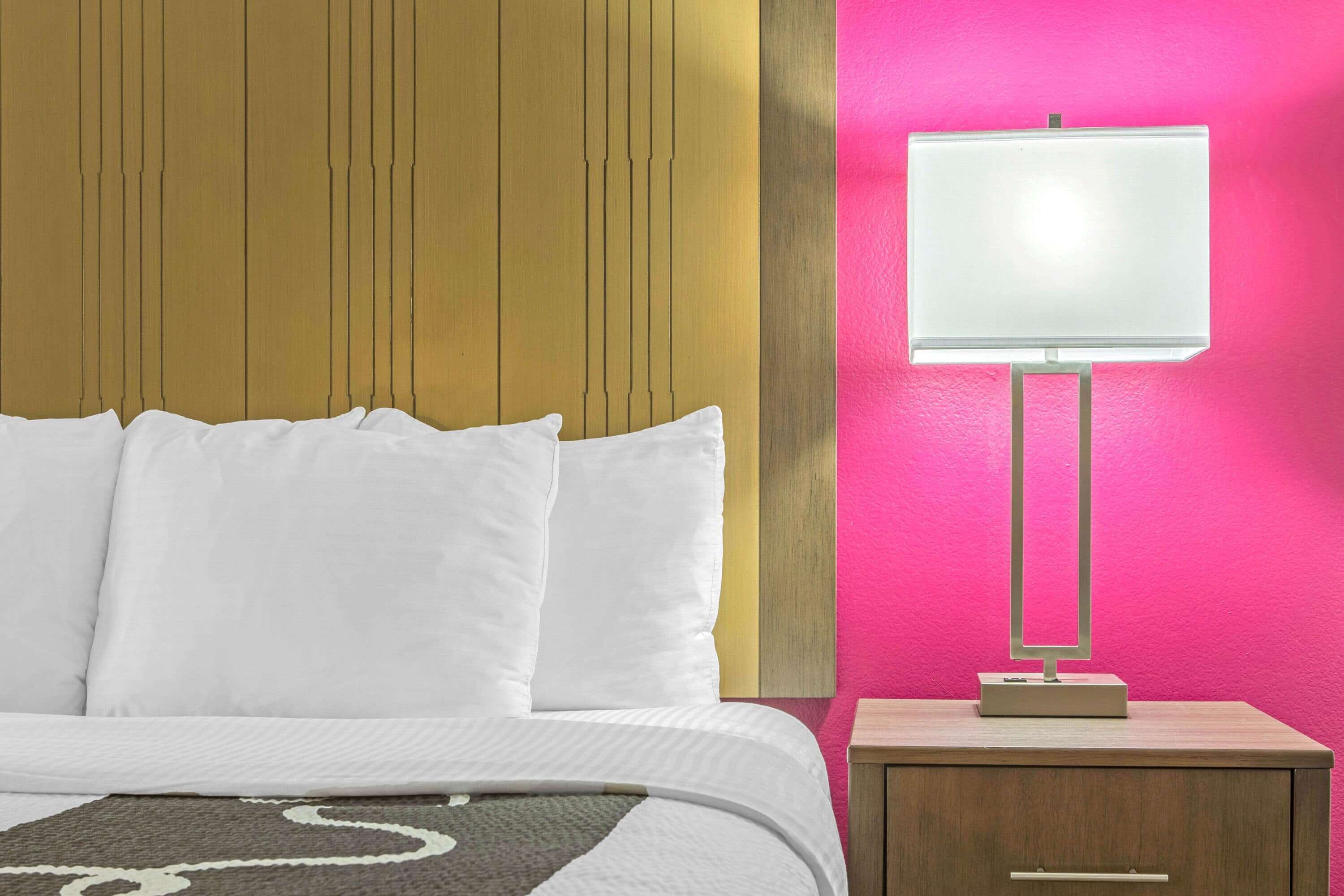 Quarto de hotel com cama, mesa de cabeceira com luminária e cabeceira de madeira.