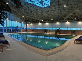 Piscina interna com paredes de vidro transparente, cadeiras de estar e plantas em vasos.
