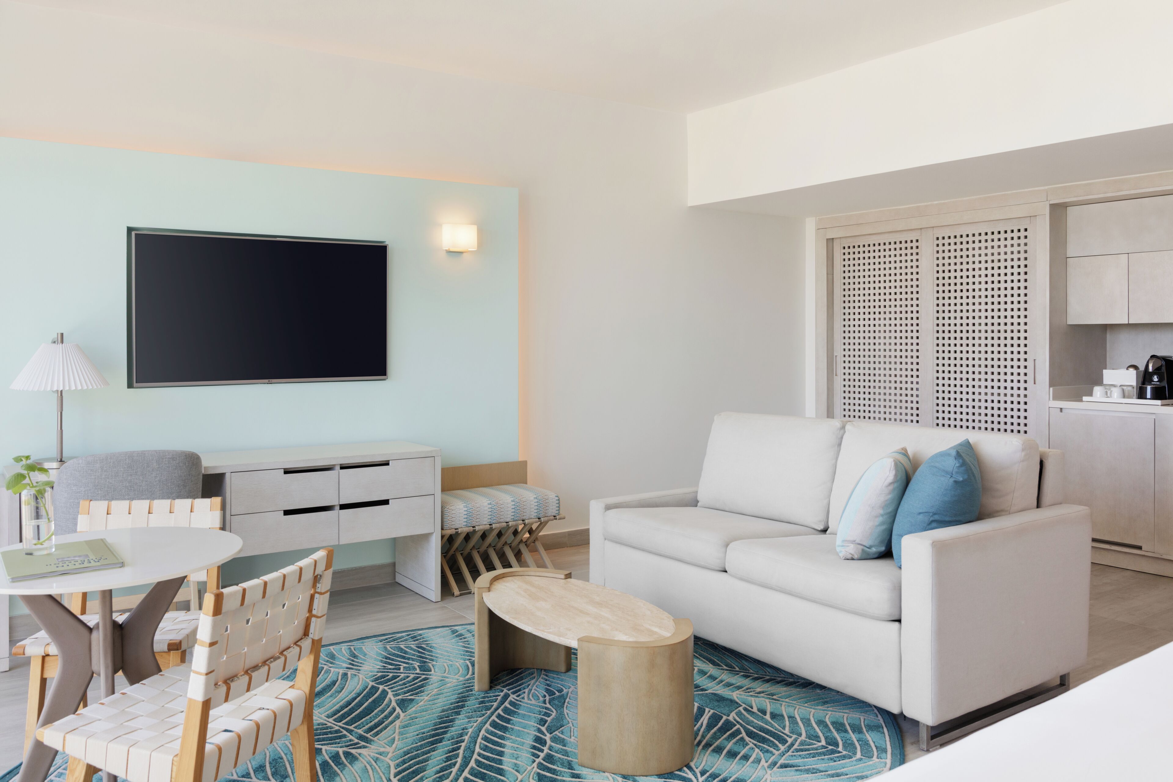 Uma sala de estar moderna com televisão de tela plana, um sofá branco com almofadas azuis, uma mesa de centro redonda e um tapete estampado.