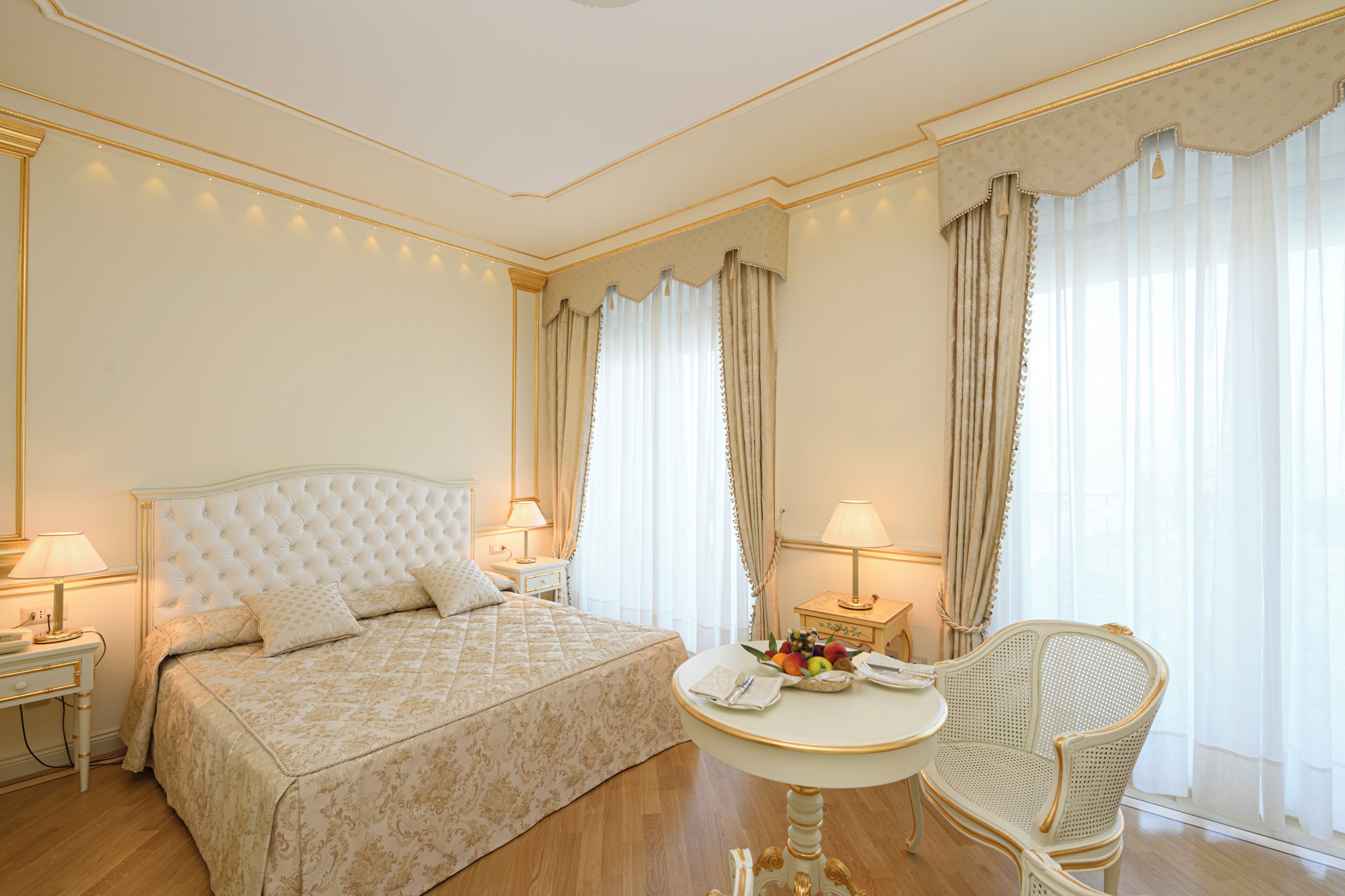 Quarto de hotel com uma cama grande, duas mesas de cabeceira com luminárias, uma mesa redonda com uma cadeira e vista para o exterior através de cortinas leves.