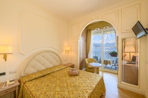 Quarto de hotel com cama, mesa de cabeceira, luminária, televisão e varanda com mesa e cadeira.