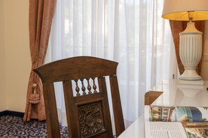 Uma cadeira de madeira com encosto entalhado e painel decorativo, uma mesa branca com abajur e uma janela com cortinas leves.