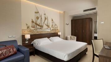 Quarto de hotel com cama, poltrona azul, guarda-roupa de madeira, escrivaninha com um laptop e um mural na parede com barcos à vela.