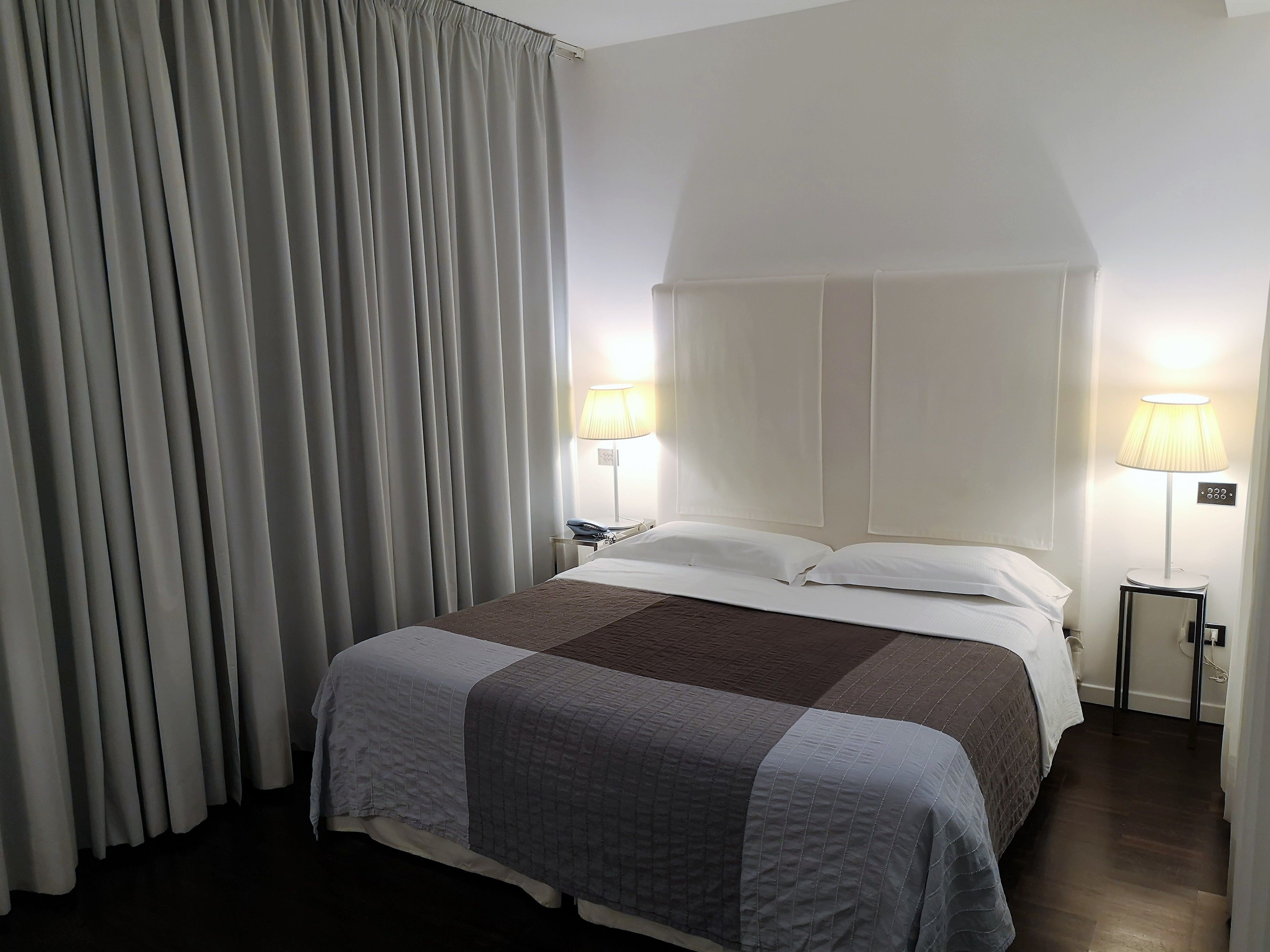 Quarto de hotel com uma cama grande, duas luminárias de cabeceira, uma mesa de apoio e uma cortina.