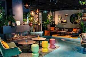 Uma área de estar moderna com lareira, mesas redondas, banquetas coloridas e diversas plantas.