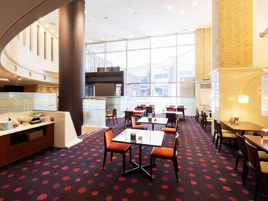 Um hall de hotel com design moderno, apresentando uma área de estar central com mesas e cadeiras, uma cozinha e grandes janelas com vista para a cidade.