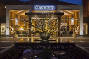 Entrada de um hotel Sheraton com um grande vaso de plantas em primeiro plano, um saguão bem iluminado e uma área de bar visível.