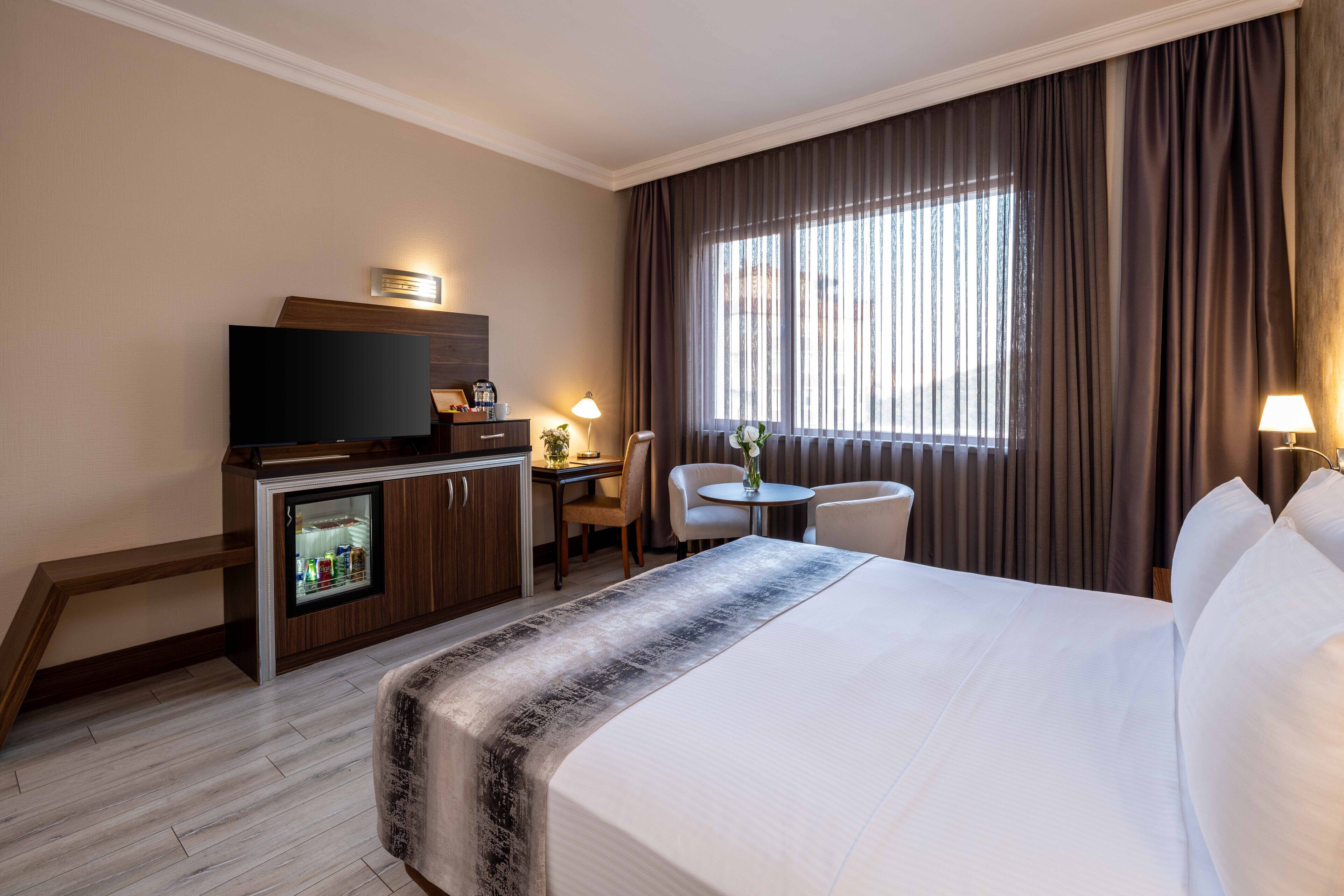 Quarto de hotel com cama, televisão, minibar, secretária com cadeira e janela com cortinas.