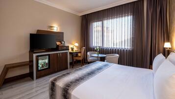 Quarto de hotel com cama, televisão, minibar, secretária com cadeira e janela com cortinas.