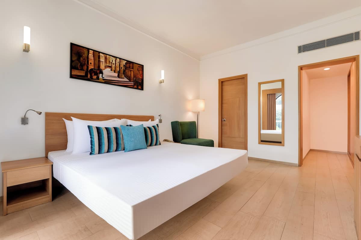 Uma cama grande com lençóis brancos, cabeceira de madeira e várias almofadas. Há uma cadeira verde, uma mesinha de cabeceira e um abajur fixado na parede. Um quadro está sobre a cama, e existem duas portas e um espelho no quarto.