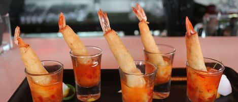 Uma bandeja com cinco copos de shot contendo uma bebida vermelha, cada um com um tempurá de camarão na borda.