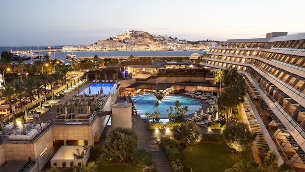 Um resort com uma piscina grande, palmeiras e vista para uma cidade histórica do outro lado da água.