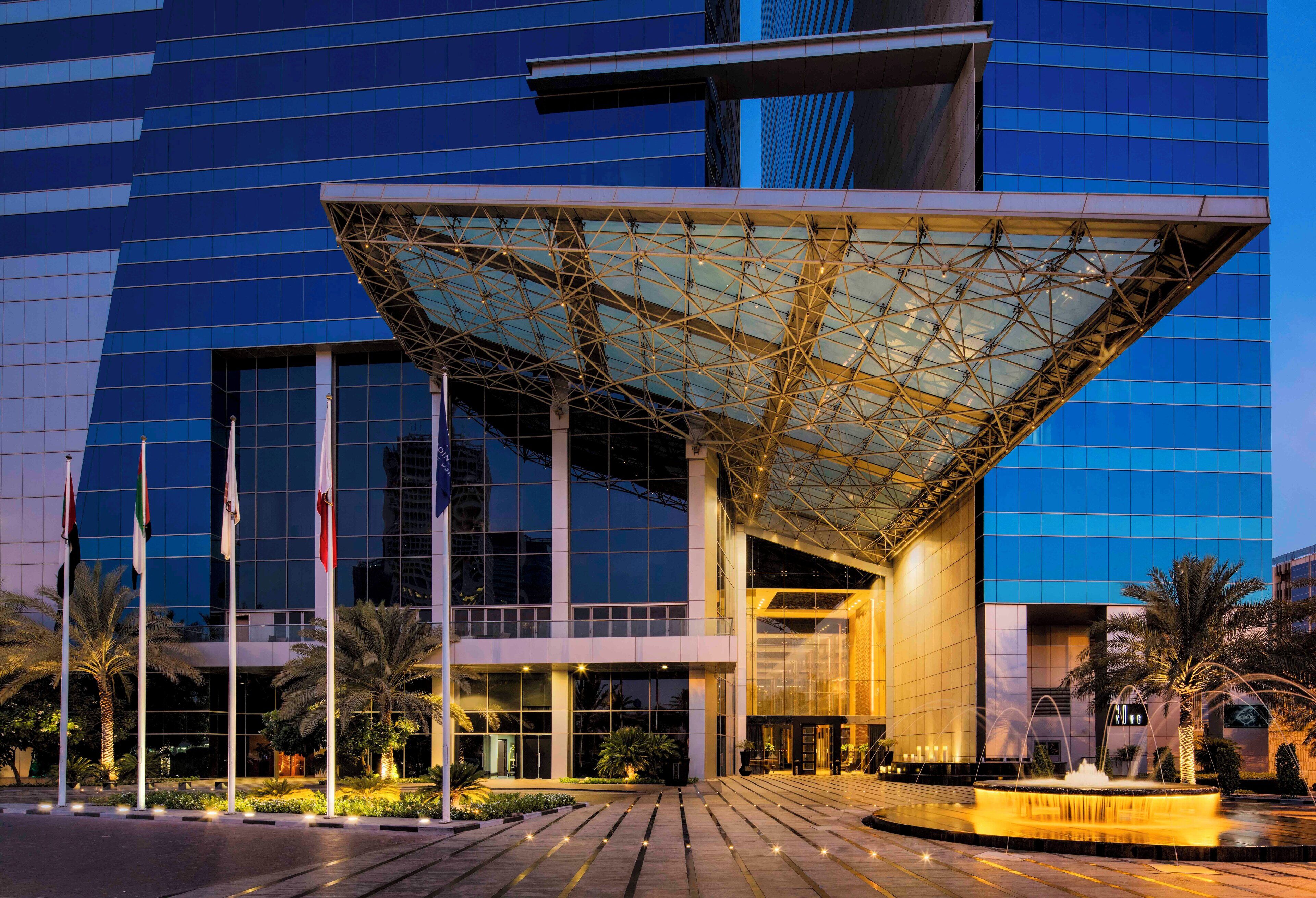 Uma entrada de hotel moderna com fachada de vidro, estrutura de dossel geométrica e um pátio ajardinado com palmeiras.