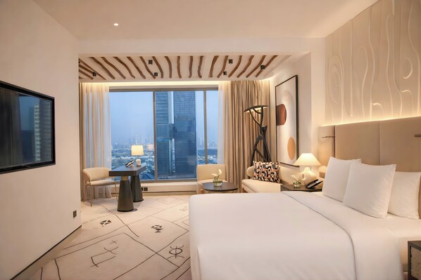 Quarto de hotel moderno com uma cama grande, uma escrivaninha, uma cadeira e vista para a paisagem urbana através de grandes janelas.