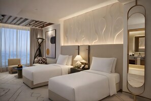 Quarto de hotel com duas camas, um espaço de estar com uma cadeira e uma pequena mesa, um espelho grande e vista da cidade através de amplas janelas.