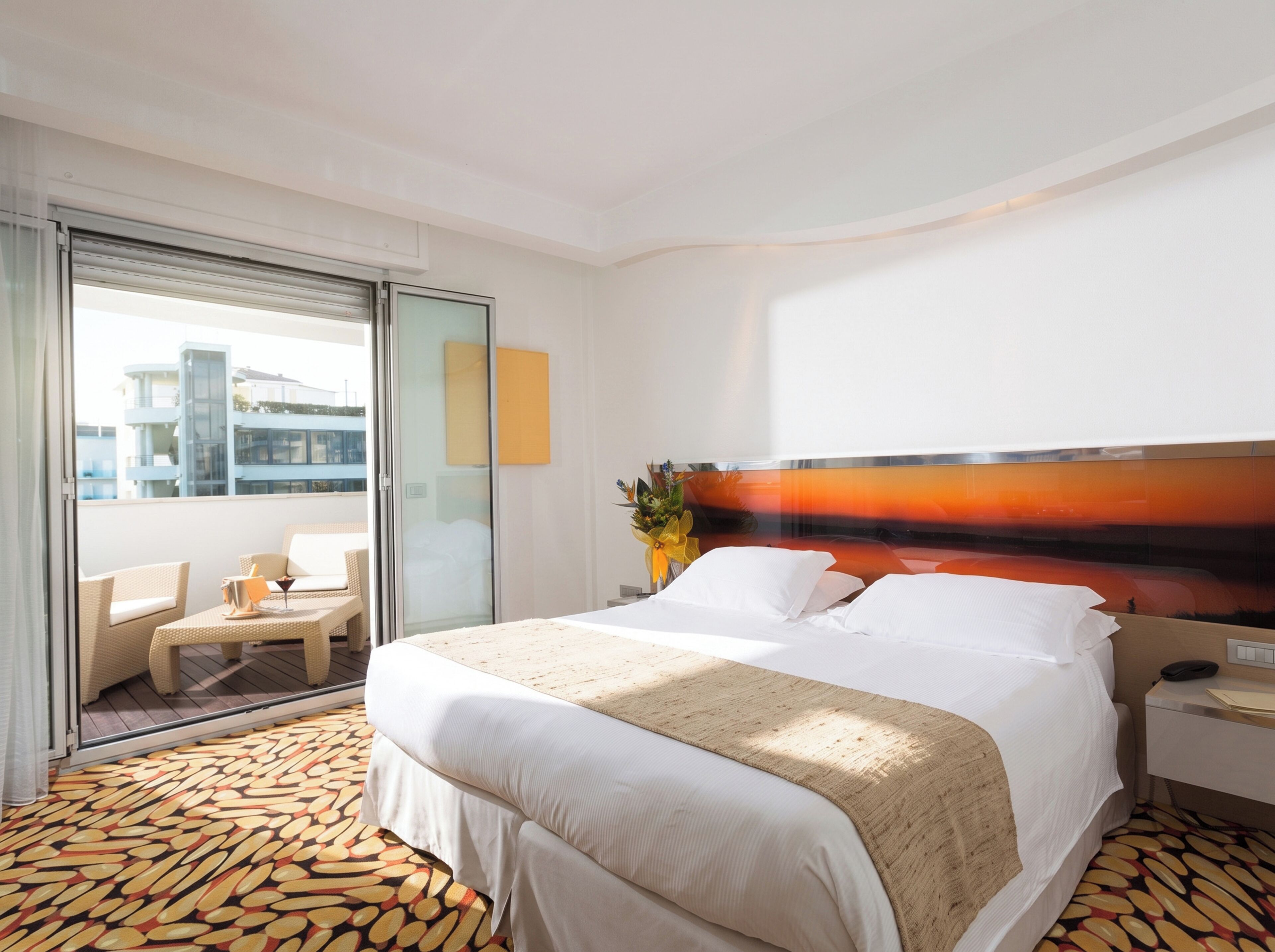 Quarto de hotel com cama, escrivaninha, cadeira, vaso de flores e vista para prédios por uma porta de vidro.