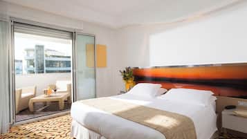 Quarto de hotel com cama, escrivaninha, cadeira, vaso de flores e vista para prédios por uma porta de vidro.