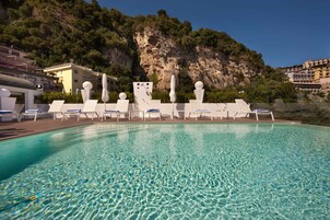 Piscina transparente com espreguiçadeiras e guarda-sóis, cercada por edifícios e uma paisagem montanhosa.