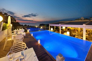 Área de piscina externa com mesas e cadeiras, um espaço coberto para sentar e vista para o mar ao entardecer.