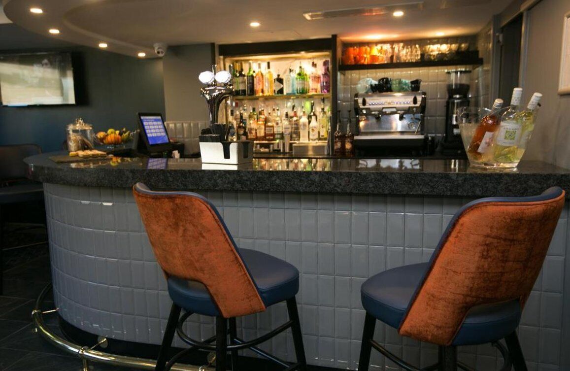 Uma área de bar com dois bancos altos, um balcão com uma tela digital e prateleiras cheias de diversas garrafas.