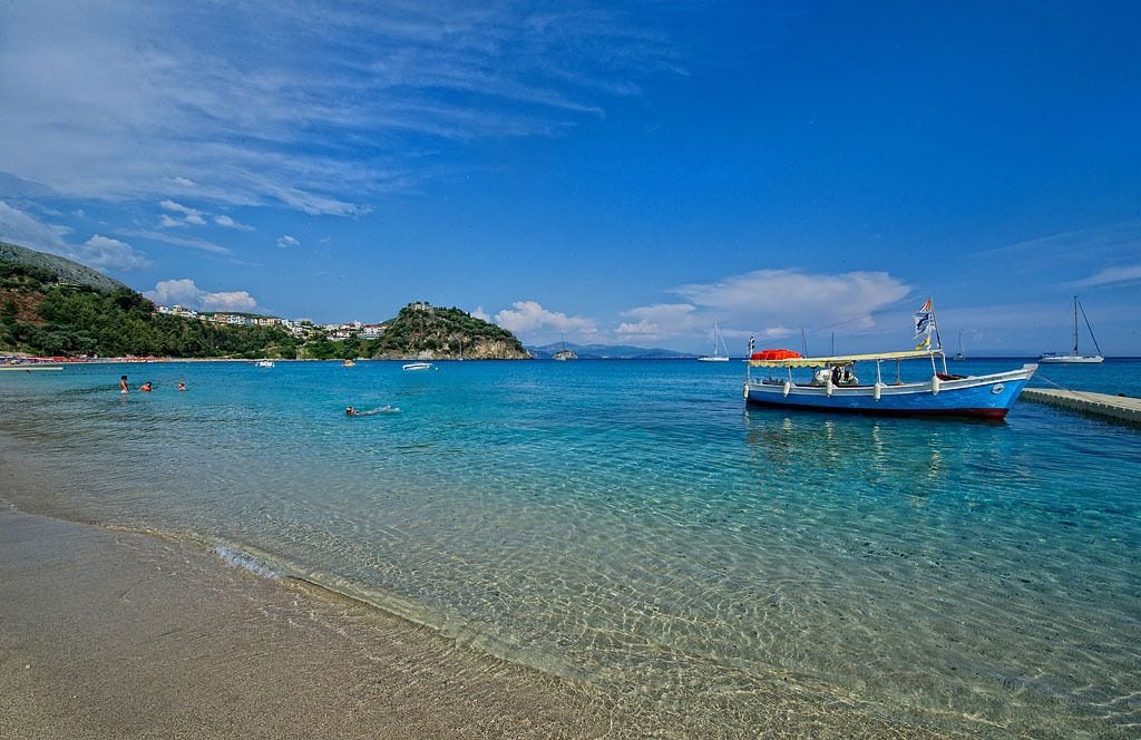 Uma praia clara com um barco pequeno atracado perto da costa, águas calmas e uma colina distante com edifícios.