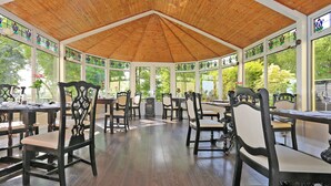 Uma área de refeições coberta com piso de madeira, cadeiras ornamentadas e grandes janelas com vitrais.