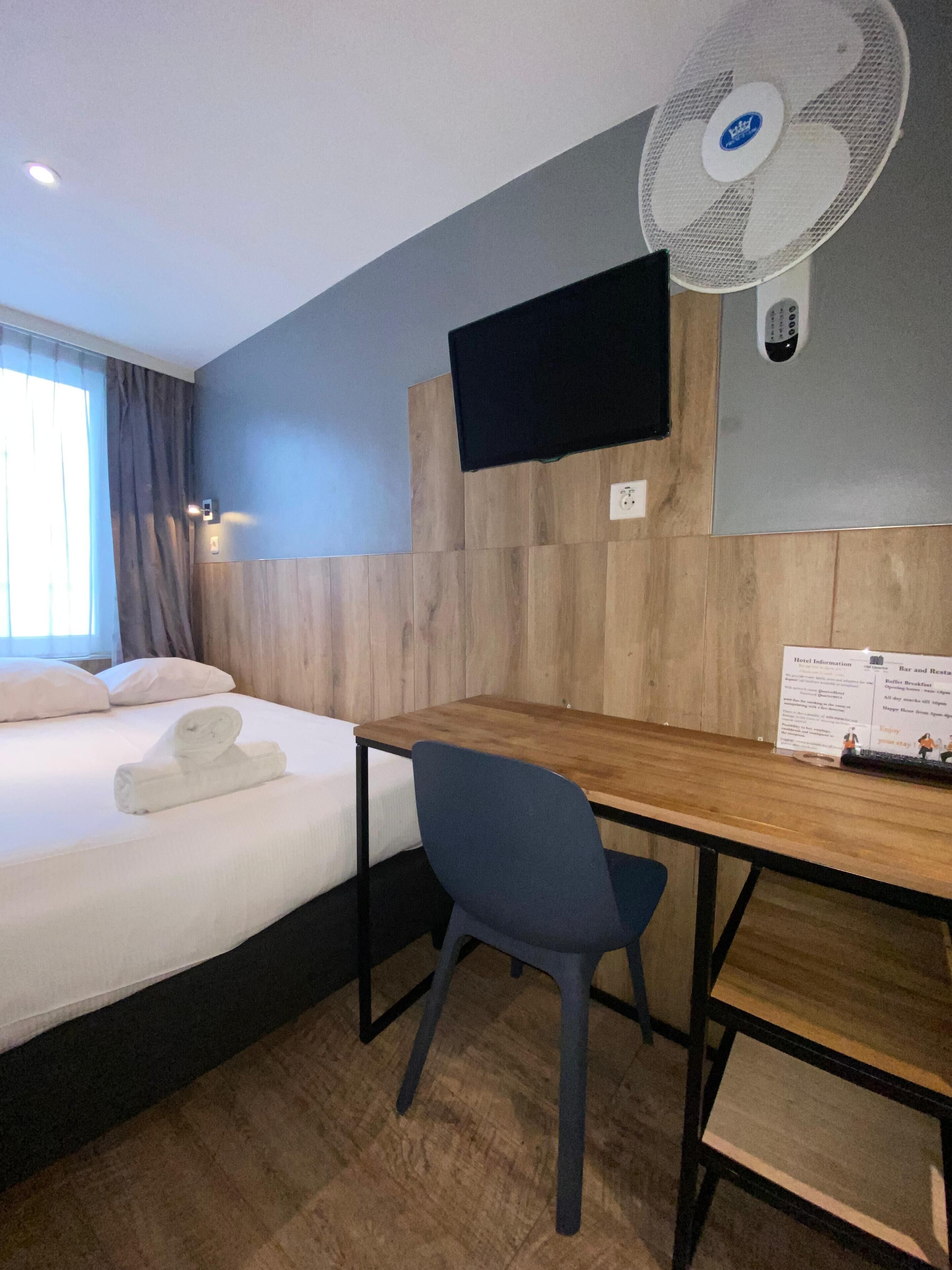 Quarto de hotel com uma cama, uma escrivaninha, uma cadeira, televisão na parede e um ventilador.