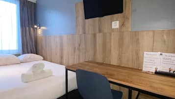 Quarto de hotel com uma cama, uma escrivaninha, uma cadeira, televisão na parede e um ventilador.